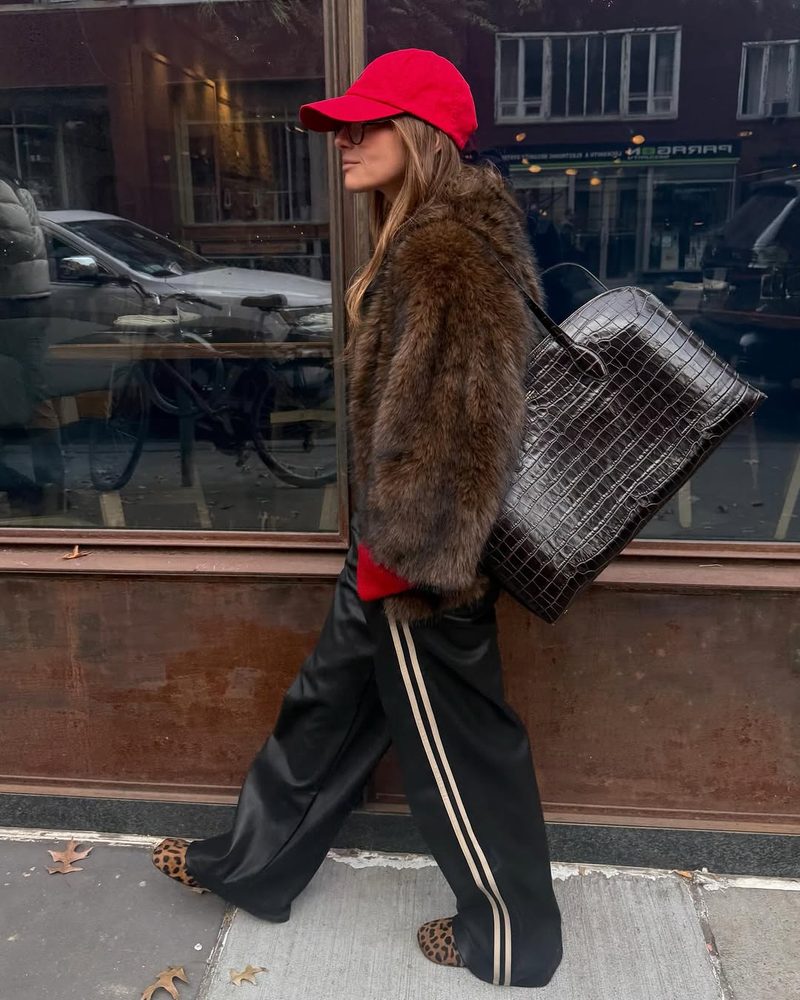 Model showcases an oversized brown fur coat paired with a bold red cap%2C black track pants with gold stripes%2C and leopard-print loafers%2C completing a chic winter look