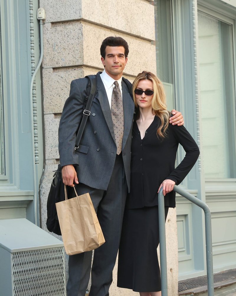 Sophisticated couple showcasing office-ready looks%3A the man in a gray pinstripe suit with a patterned tie%2C the woman in a chic black blouse and skirt with stylish sunglasses.