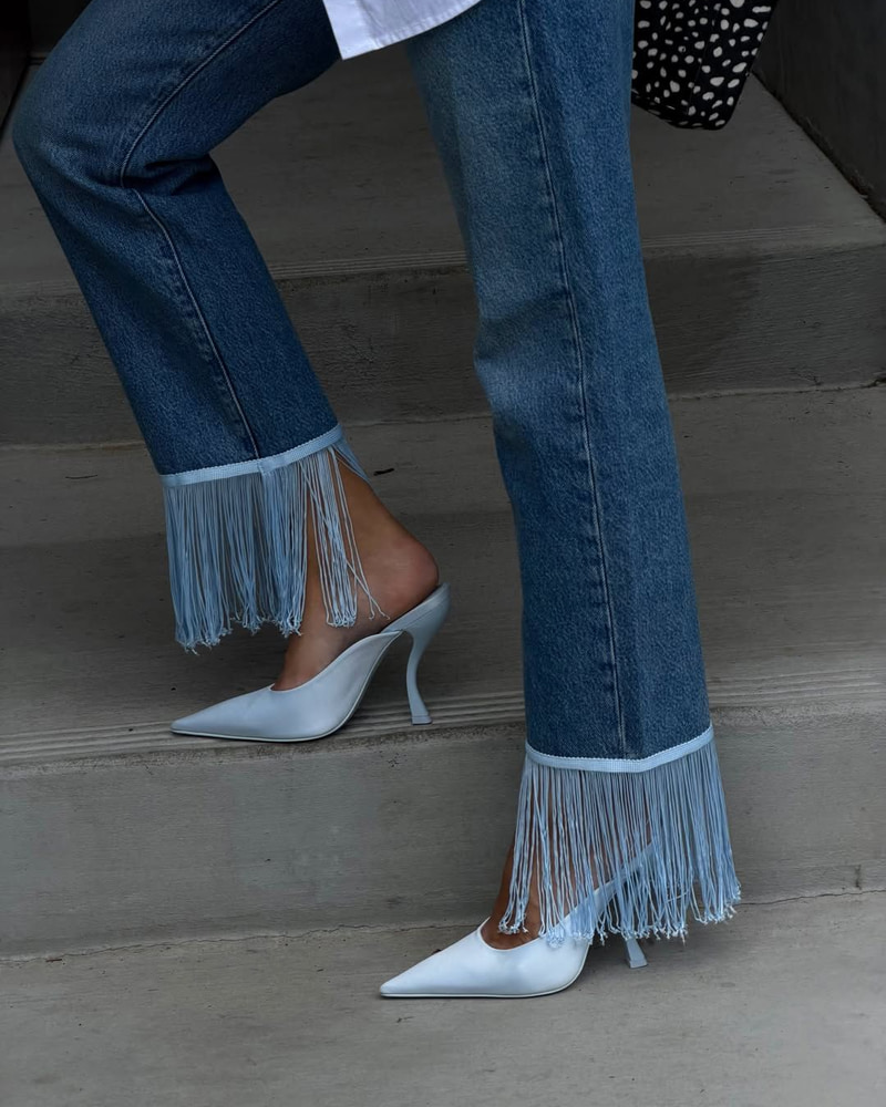 High-waisted denim jeans feature playful fringe detailing at the hem%2C styled with sleek white pointed-toe heels for a chic look