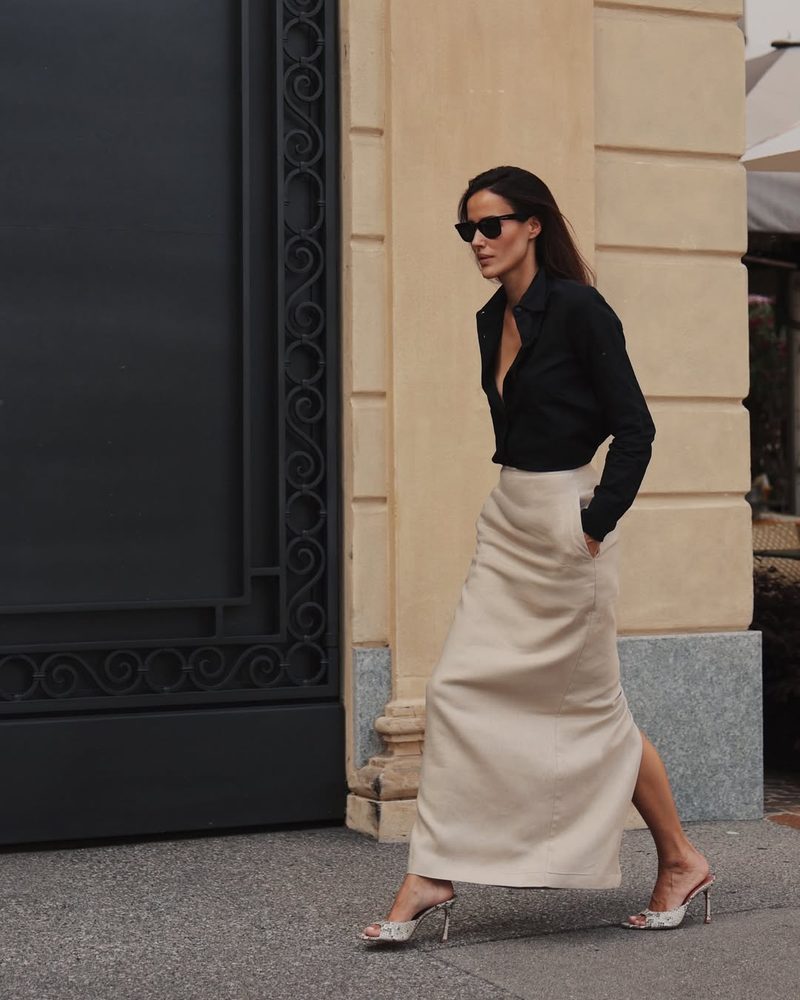 Street style look featuring a black button-down shirt tucked into a beige maxi skirt with slit%2C styled with heels on a city sidewalk