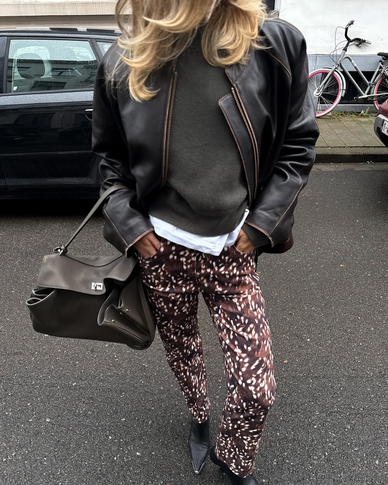 Chic layered outfit featuring a brown leather jacket%2C olive green sweater%2C patterned brown denim pants%2C and sleek black ankle boots