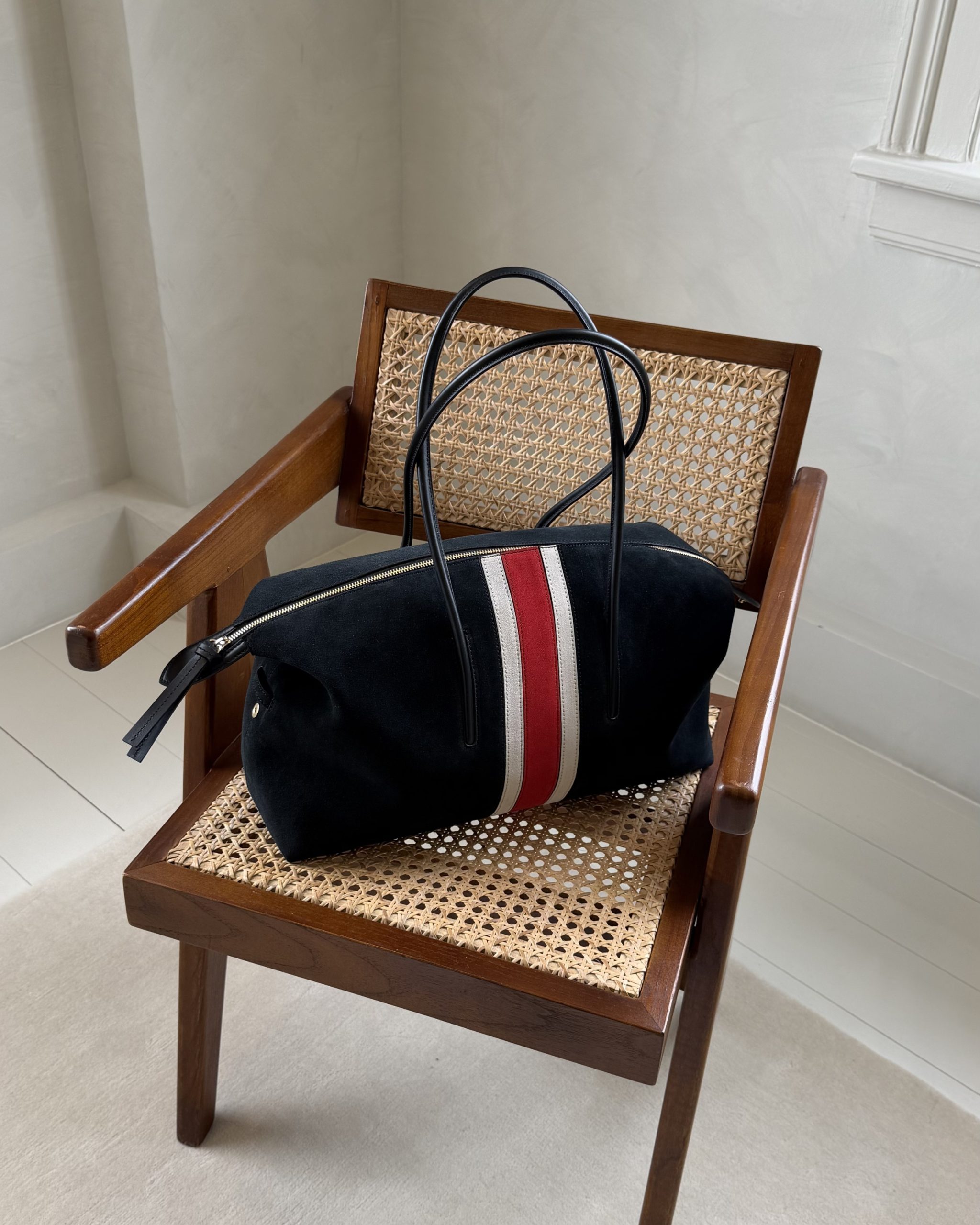 Black suede tote bag with red and white vertical stripes and leather handles on a wooden cane chair.