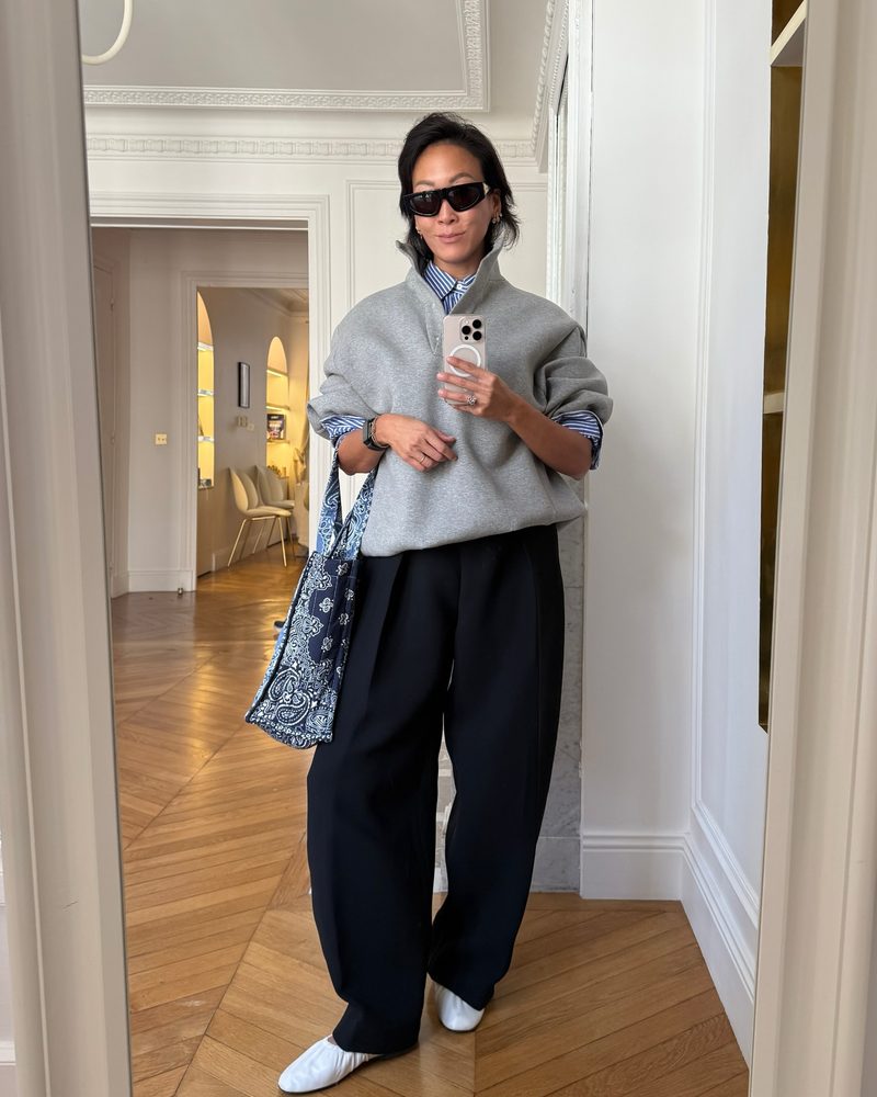 Relaxed gray quarter-zip sweater layered over a blue striped shirt%2C paired with black wide-leg trousers and white slip-on shoes%2C complemented by a patterned tote bag