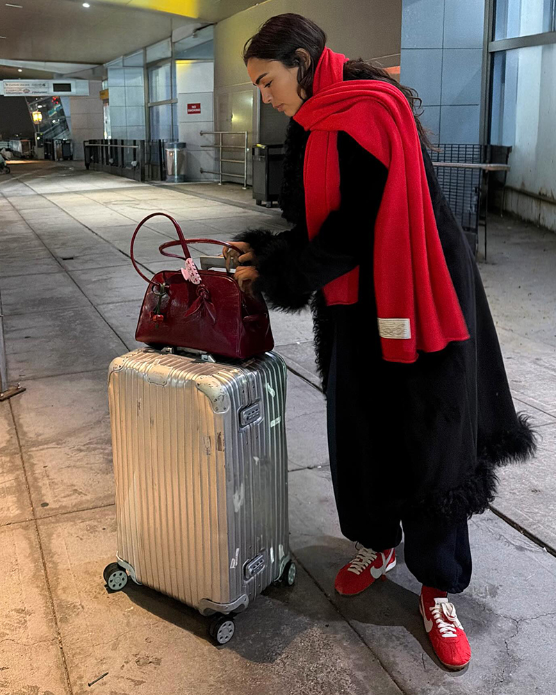 Travel style moment featuring black shearling coat%2C red scarf and sneakers%2C styled with leather tote at airport curbside