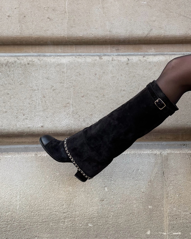 Close-up of black suede knee-high boots with chain detail and buckle%2C styled with sheer tights on stone steps