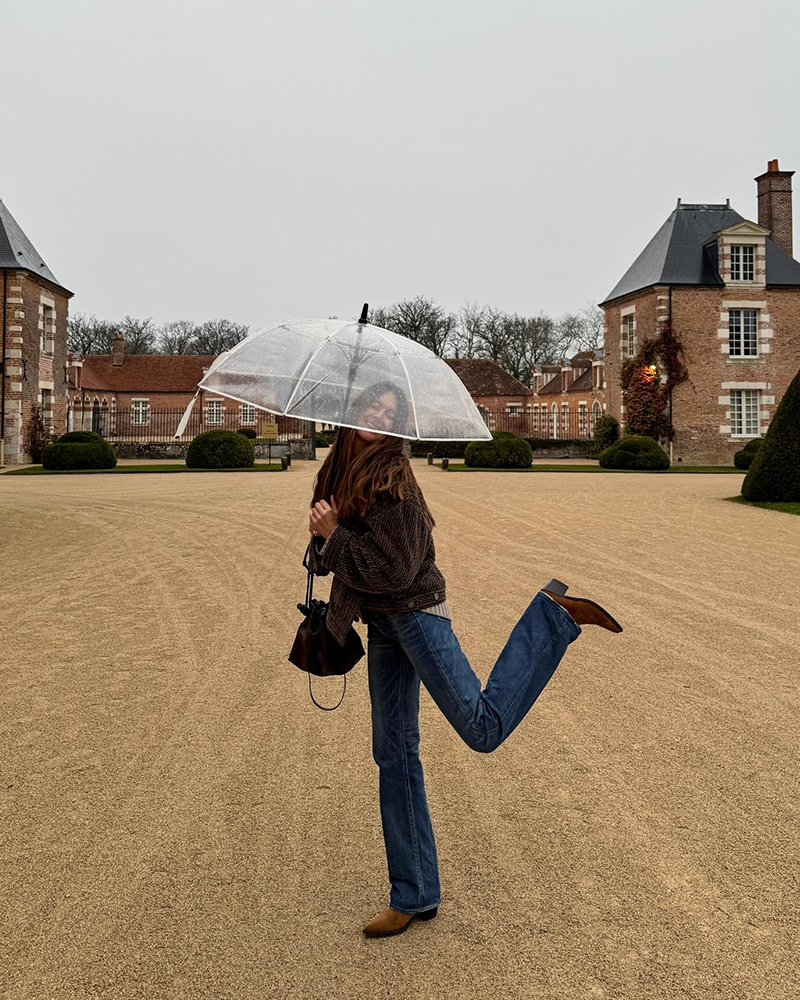 Woman in straight-leg blue jeans%2C brown leather boots%2C and knit sweater holding clear umbrella in courtyard of historic estate