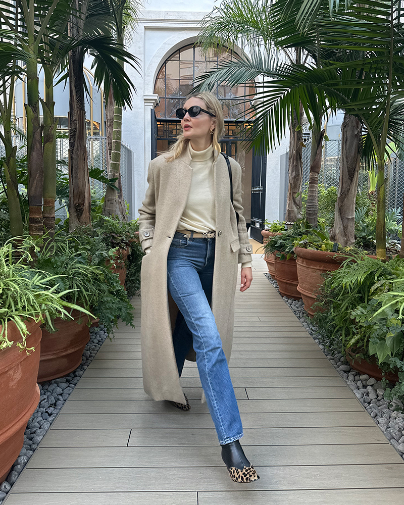 Street style look in a beige wool coat%2C cream turtleneck%2C blue jeans%2C leopard ankle boots%2C walking through a palm-lined courtyard