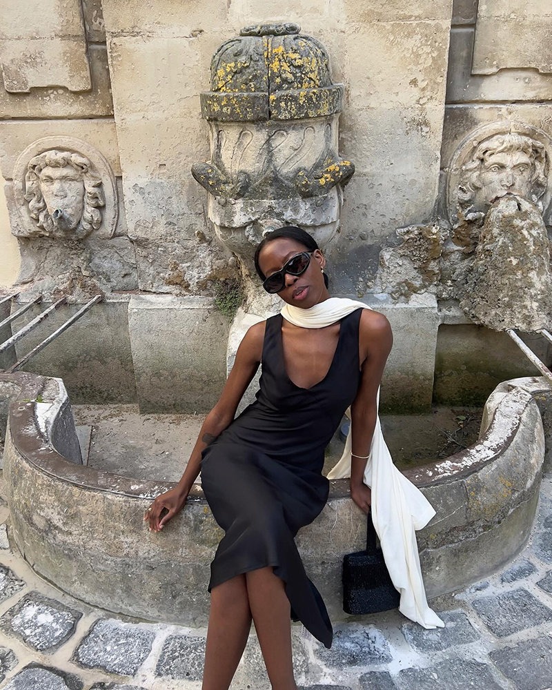 Black satin slip dress with a cream scarf%2C cat-eye sunglasses and clutch%2C styled at a historic stone fountain courtyard