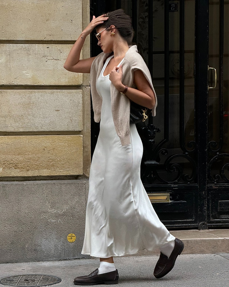 Ivory satin midi slip dress with beige knit draped over shoulders and brown loafers on a Paris street