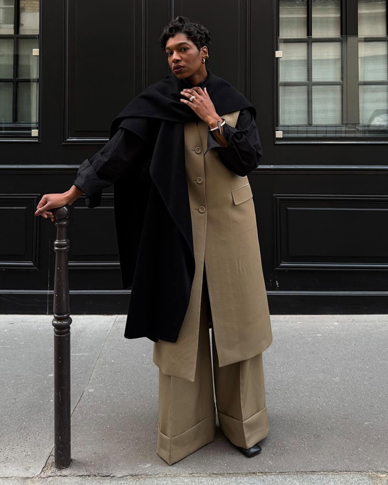 Layered fall outfit with a camel longline vest%2C wide-leg trousers and black draped scarf on a city street%0A