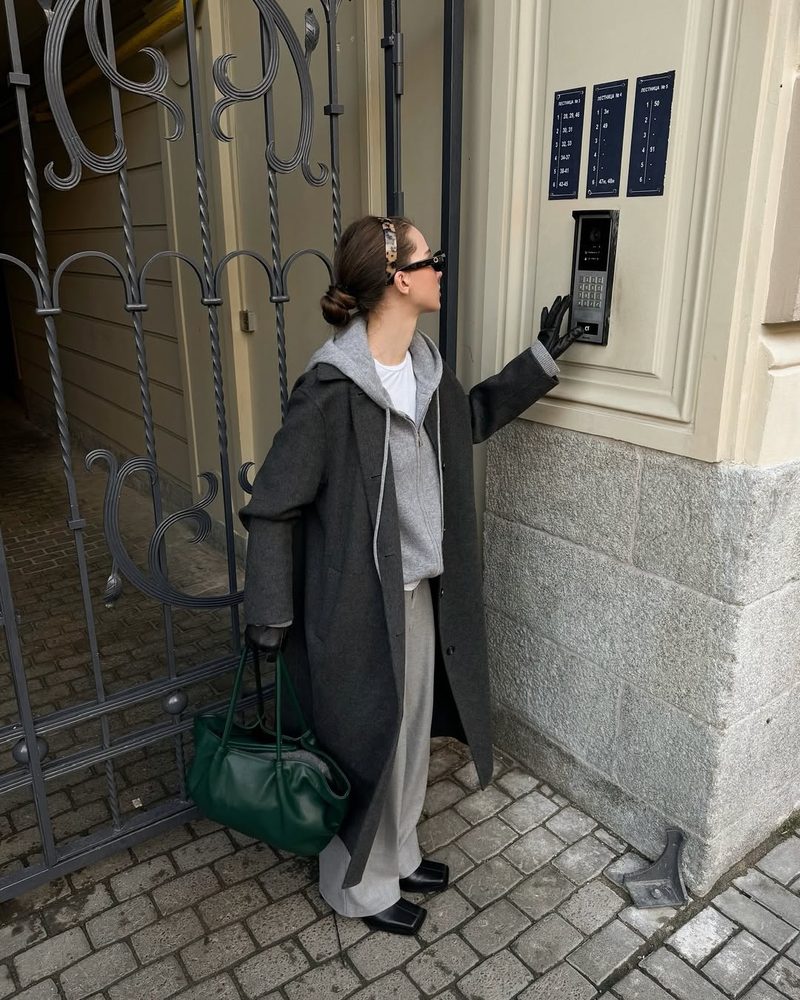 Chic winter outfit featuring a dark overcoat%2C gray hoodie%2C and relaxed trousers%2C complemented by a green leather handbag and sleek sunglasses.