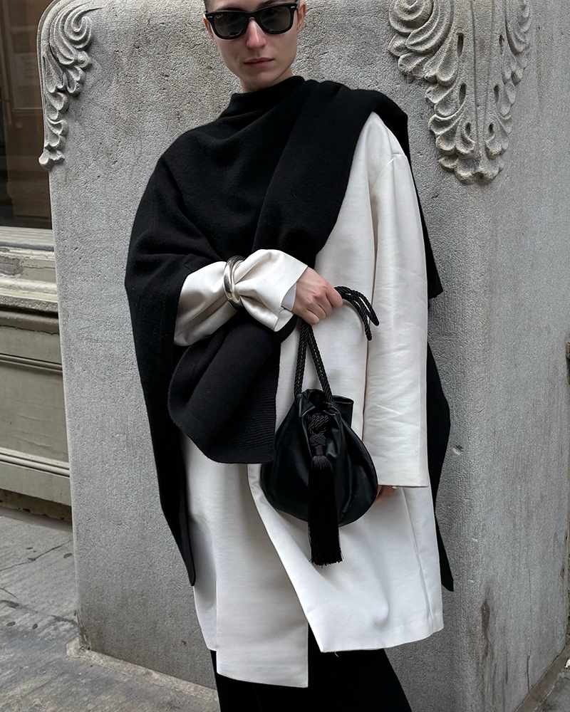 Black and white minimalist outfit with oversized cape coat%2C leather bucket bag and sunglasses against stone facade