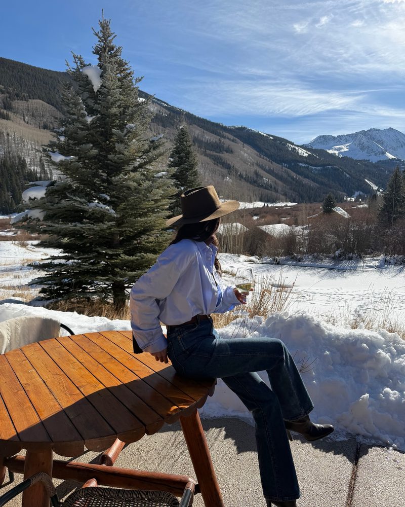 Casual mountain vacation outfit featuring a white oversized button-up shirt and dark high-waisted jeans paired with chic black ankle boots. A wide-brimmed brown hat completes the stylish look against a stunning snowy mountain backdrop