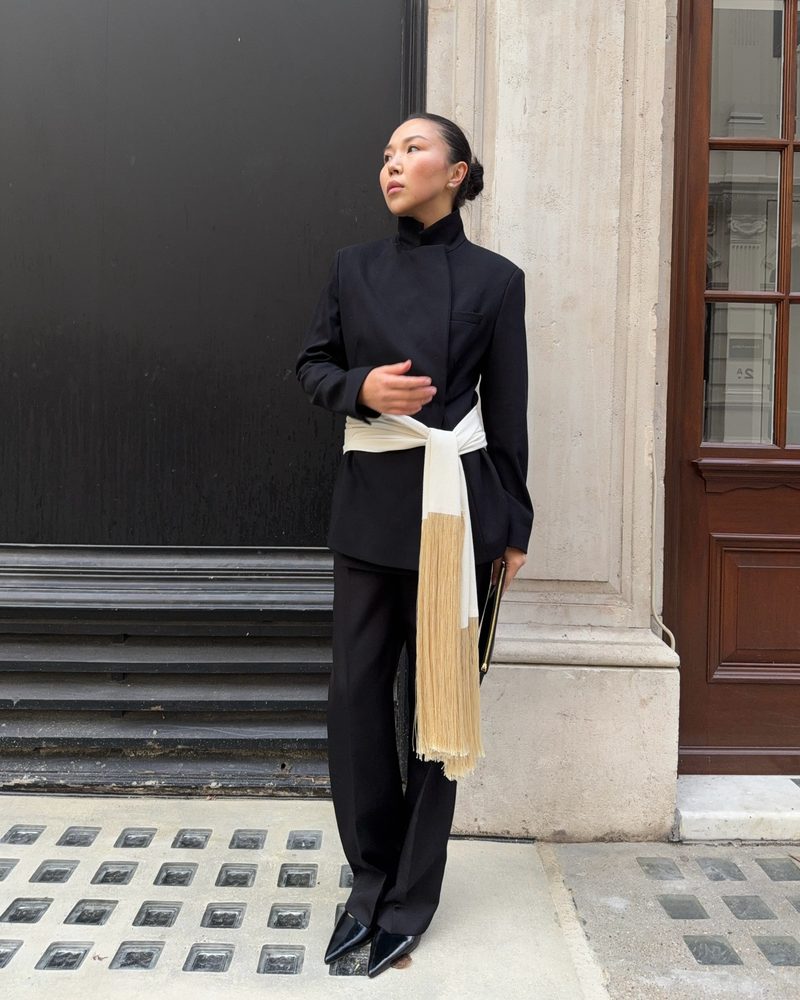 Chic black tailored blazer with a high collar%2C styled with a cream fringe belt and sleek black trousers%2C accentuated by pointed heels