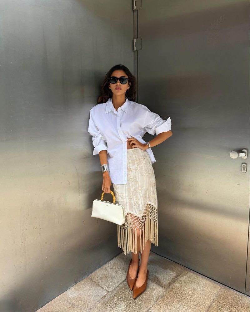 A woman poses confidently against a metallic door%2C wearing a white blouse and a cream skirt with fringe. She holds a white handbag and wears sunglasses and brown heels.