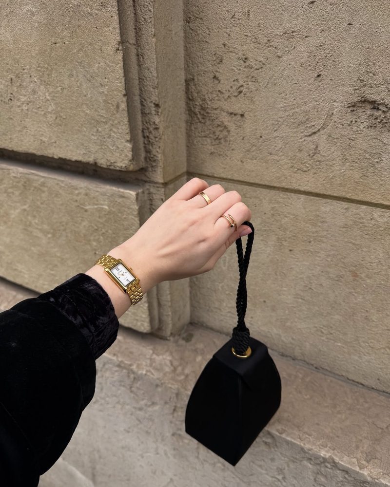 Elegant black leather handbag with a unique silhouette%2C paired with a vintage gold watch and stacked rings against a textured wall