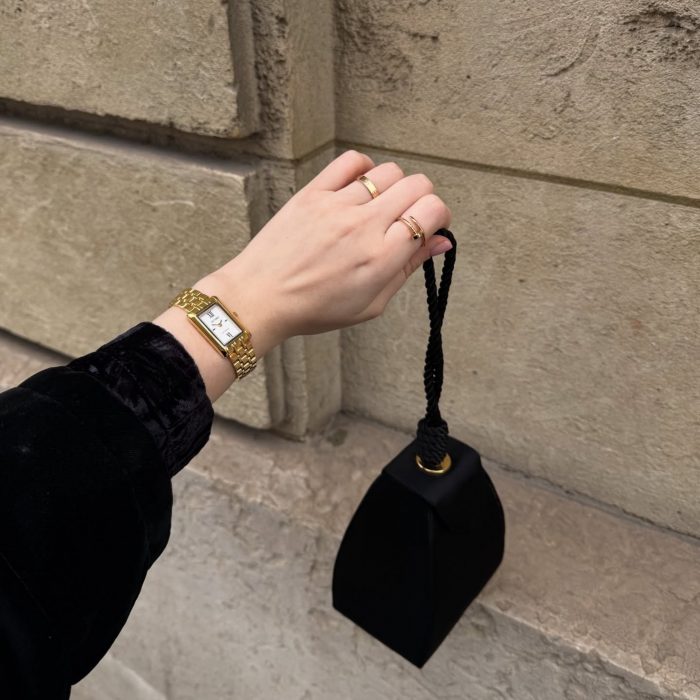 Elegant black leather handbag with a unique silhouette, paired with a vintage gold watch and stacked rings against a textured wall