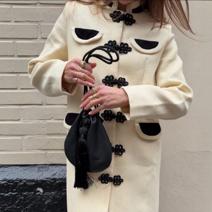 Chic cream coat with black accents features decorative buttons and pockets, paired with a sleek black satin pouch bag with a stylish tassel