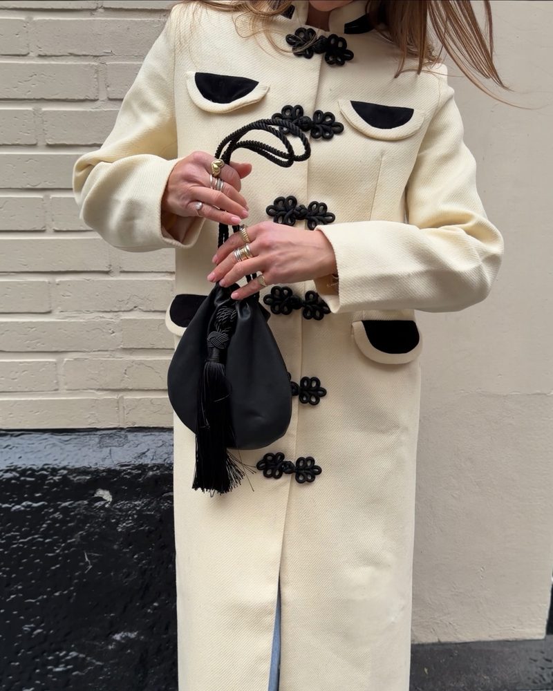 Chic cream coat with black accents features decorative buttons and pockets%2C paired with a sleek black satin pouch bag with a stylish tassel