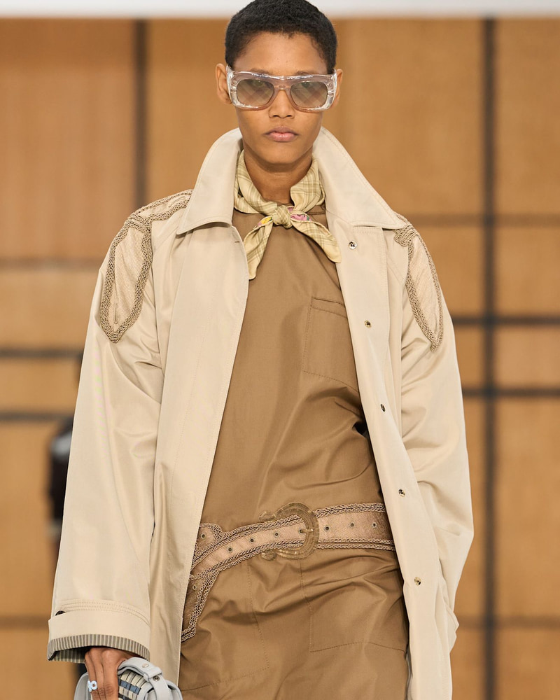 Model wearing Burberry spring trench coat over tan belted dress with check scarf and oversized sunglasses%2C holding a gray check handbag