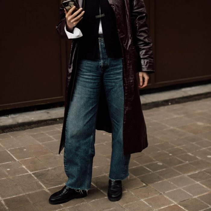 Person wearing a long brown leather coat, black sweater, and blue jeans stands on a tiled pavement, holding a cellphone, conveying a casual, stylish vibe.