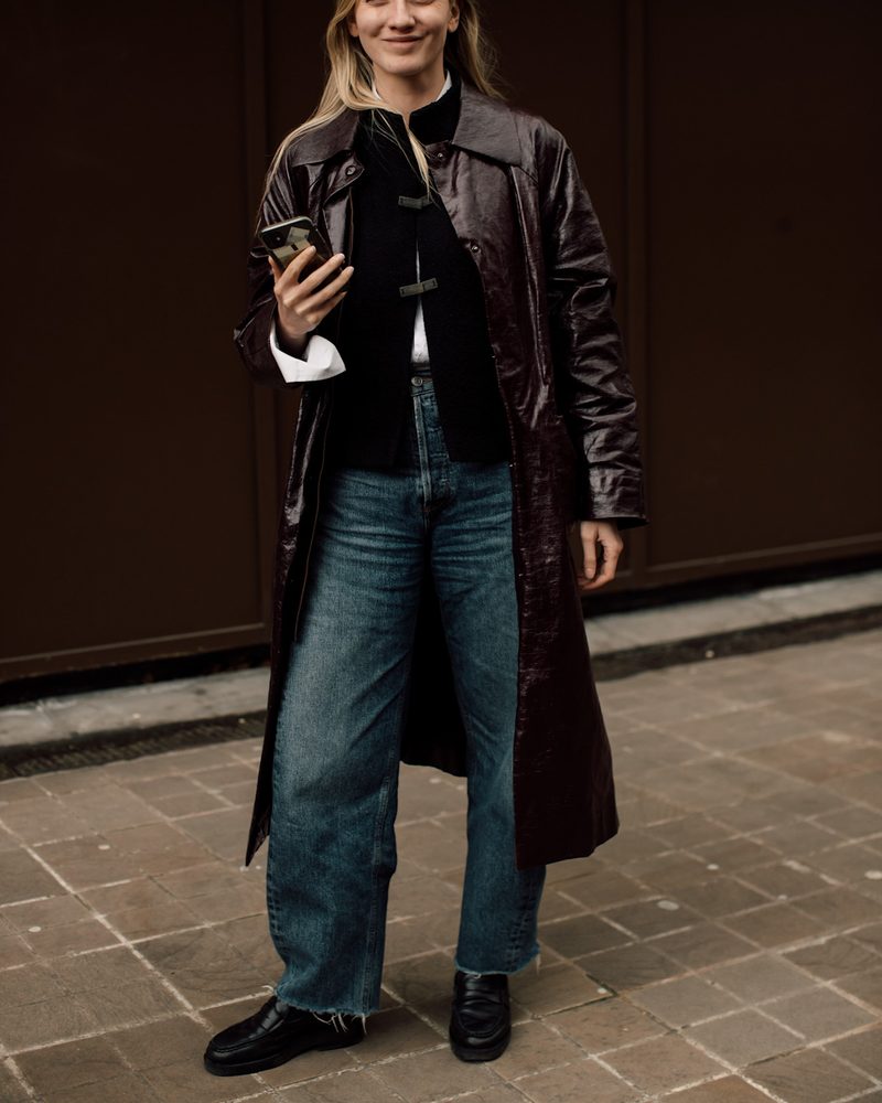 Burgundy patent leather trench coat layered over a black knit sweater%2C paired with high-waisted blue denim jeans and black loafers at London Fashion Week