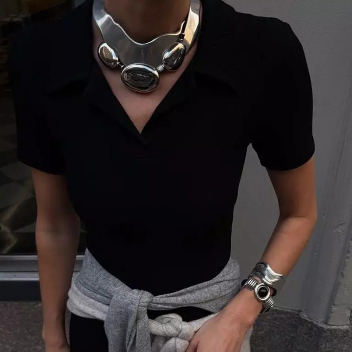 Statement silver collar necklace styled with a black V-neck top and silver cuff bracelet, layered with sweater at waist against an urban wall