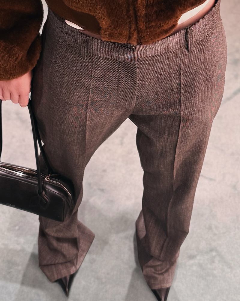 Tailored brown wool trousers styled with pointed leather boots and a mini black handbag%2C photographed on a polished concrete floor