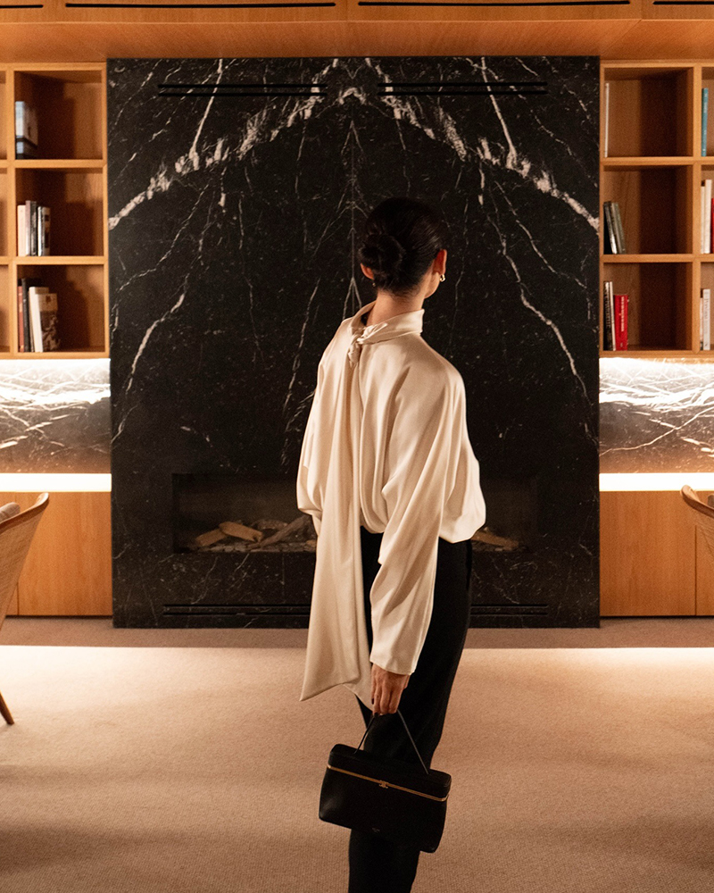 Woman in ivory draped blouse and black trousers holding a structured top-handle bag in a modern marble lounge
