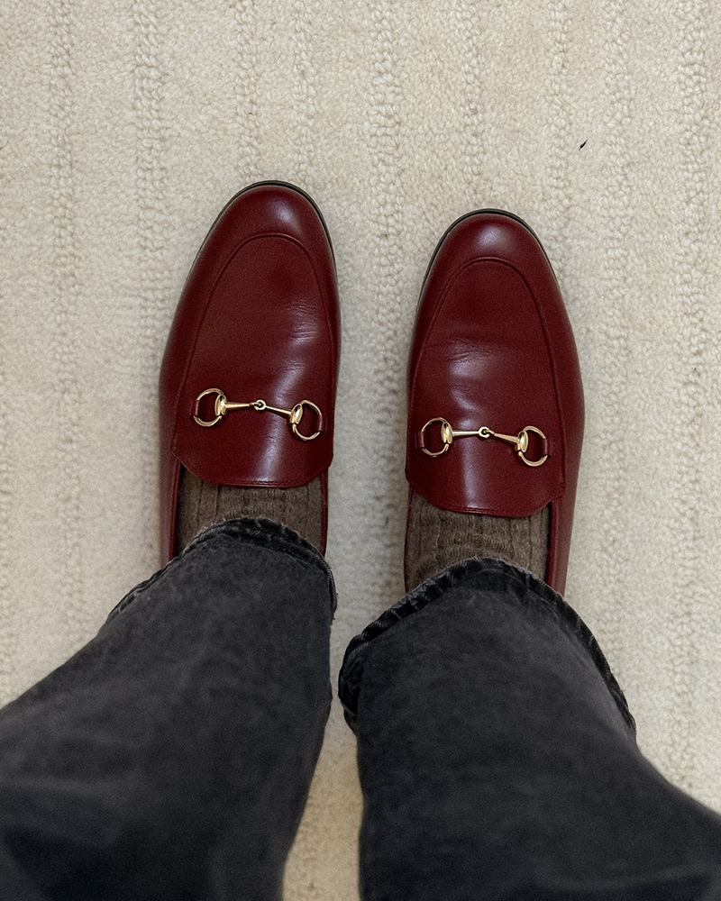 Brown loafers with gold buckle detailing are worn over brown socks and gray jeans on a light textured carpet. The style is polished and classic.