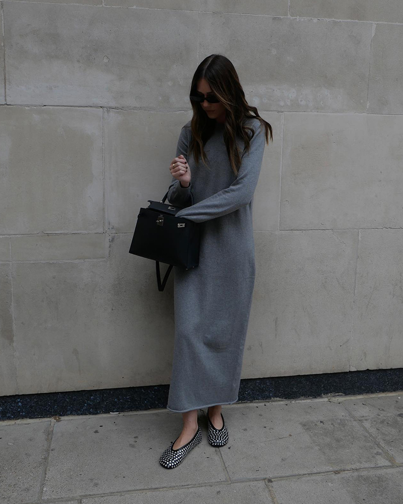 Woman in gray long-sleeve knit maxi dress with black leather top-handle bag and studded flats against stone wall