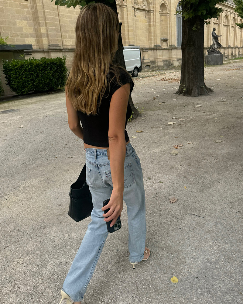 Back-view street style look with black fitted tee%2C light-wash low-rise jeans%2C black shoulder bag%2C strolling through tree-lined courtyard
