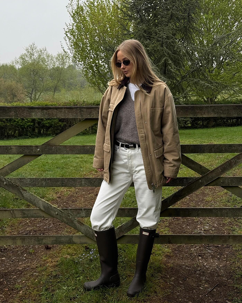 Tan utility jacket styled with white trousers and black knee-high rain boots beside a wooden fence in the countryside