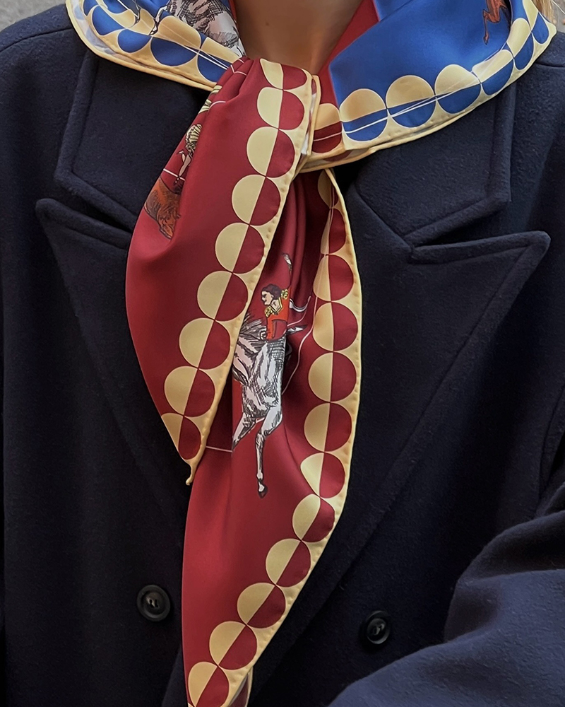 Close-up of red silk scarf with equestrian print and geometric border%2C styled over navy wool coat