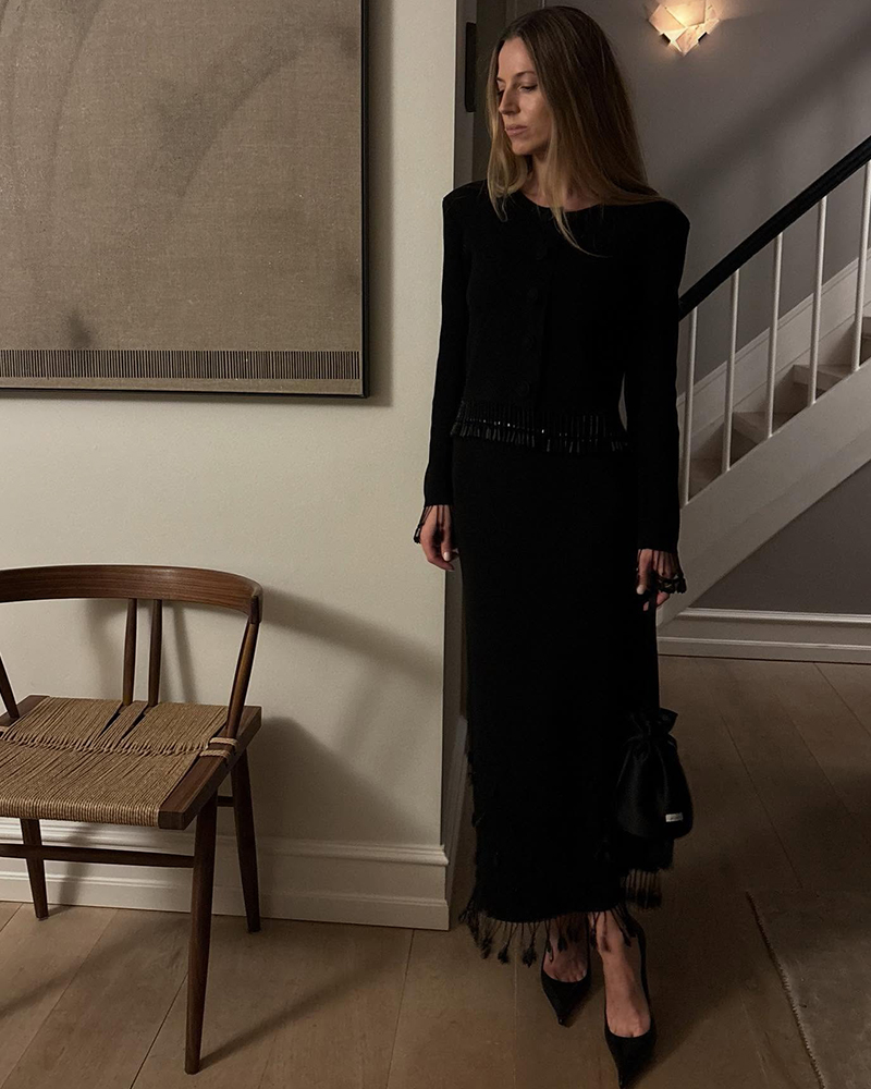 Minimalist black knit midi dress with fringe hem and pointed heels%2C styled indoors beside a wooden chair and staircase