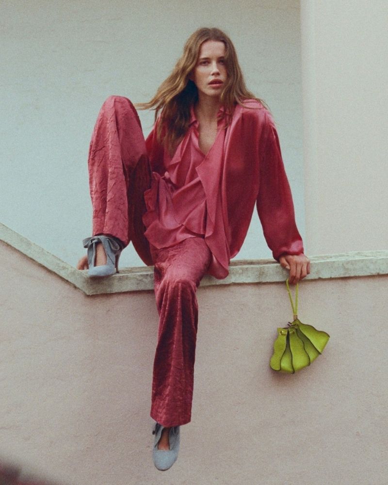 Model in rose satin blouse and velvet trousers with grey heels and green mini bag on rooftop ledge
