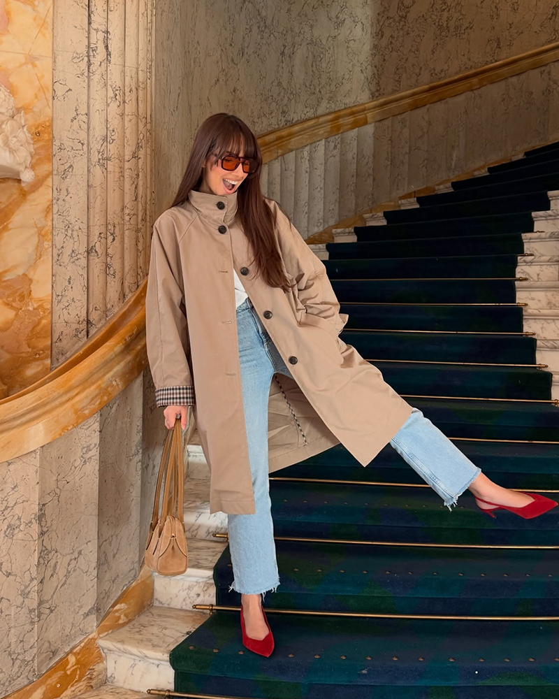 Woman in beige trench coat with cropped jeans%2C red heels and tote bag%2C posing on marble staircase indoors