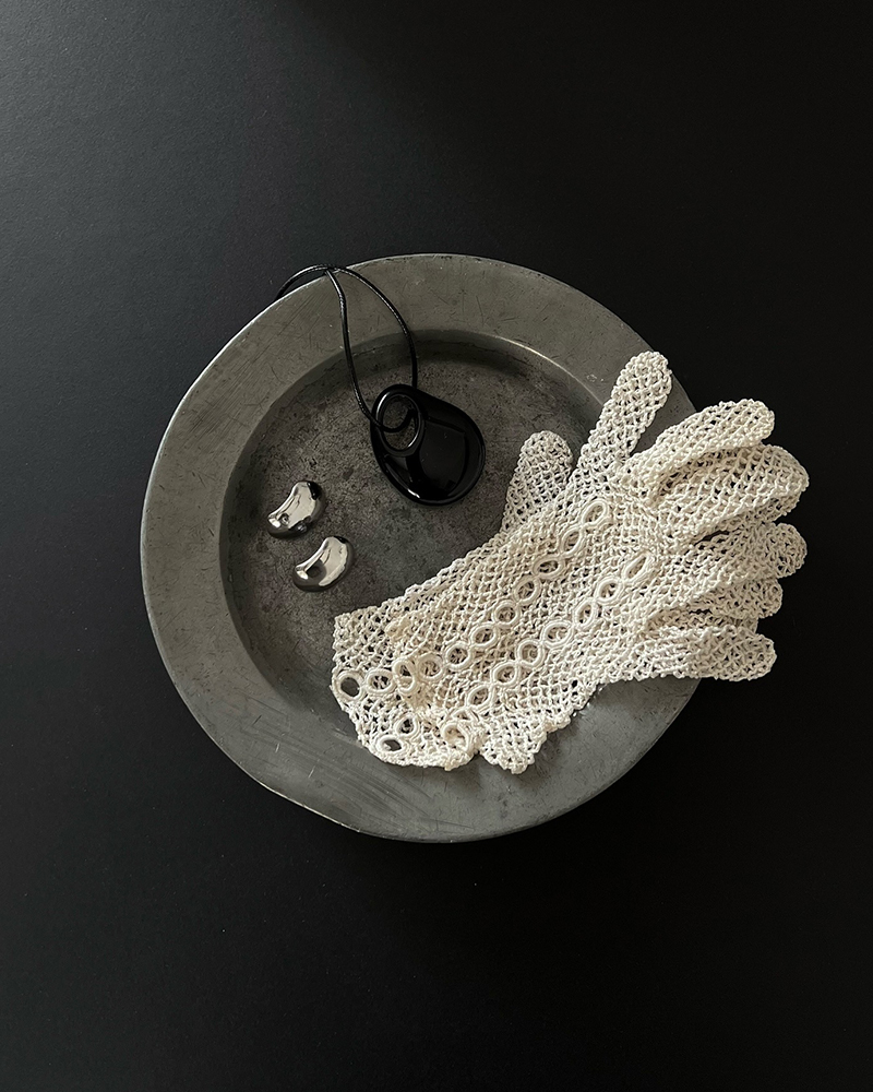 Vintage lace gloves with silver earrings and black pendant necklace styled on metal tray against dark tabletop background