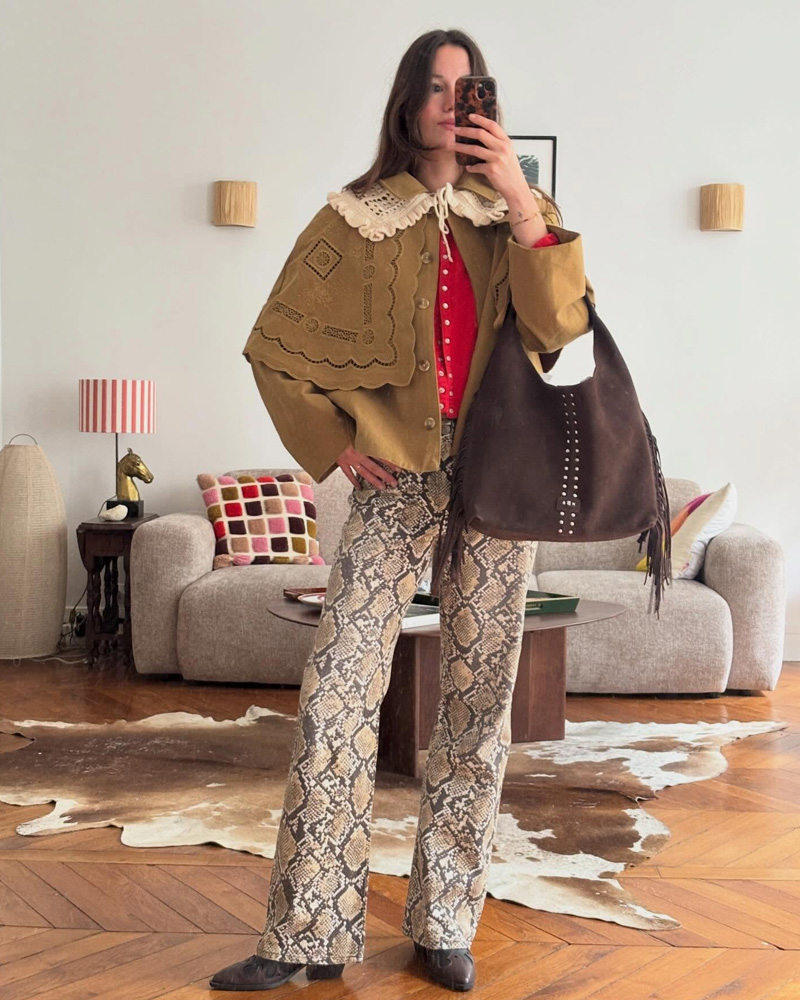Woman in embroidered cropped jacket and snakeskin flared pants holding brown leather hobo bag in chic living room