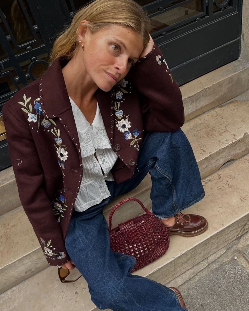 Embroidered burgundy floral jacket with white ruffle blouse%2C straight-leg jeans and woven handbag on city steps
