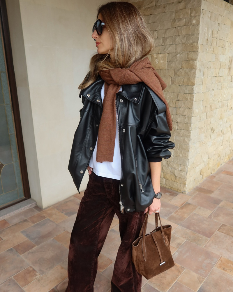 Street style look with black leather jacket%2C brown scarf%2C velvet trousers%2C and suede tote bag%2C styled with sunglasses by stone wall backdrop