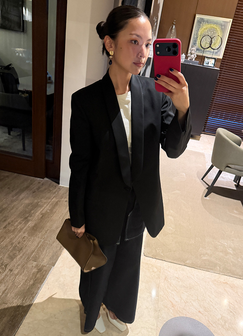 Woman in black tailored blazer and midi skirt holding brown leather clutch in modern home interior