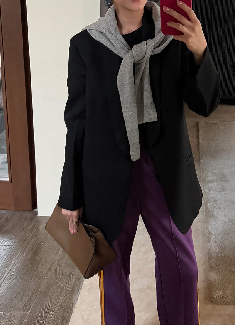Woman in black oversized blazer with grey knit scarf and purple tailored trousers holding brown leather clutch indoors