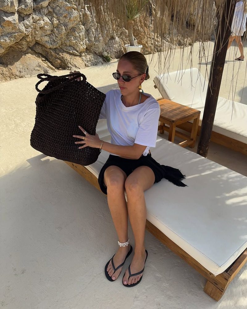 Woman wearing a white cotton t-shirt%2C black wrap skirt%2C black flip-flops%2C and holding a large black woven tote bag under a beach umbrella.