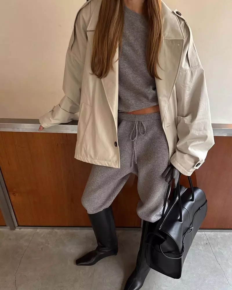 Chic gray sweatpants paired with a cropped sweater and oversized cream trench coat%2C complemented by sleek black knee-high boots and a structured tote.