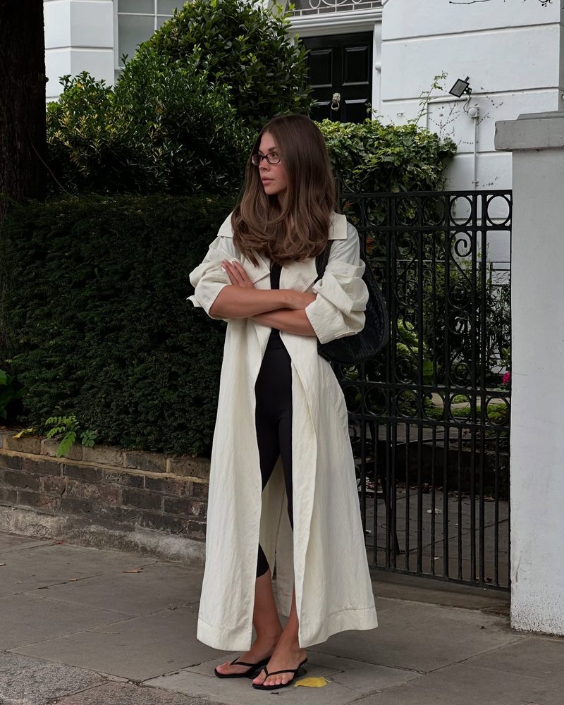 Flowy cream duster coat paired with sleek black leggings and minimalist flip-flops%2C set against a lush green backdrop for a chic spring look
