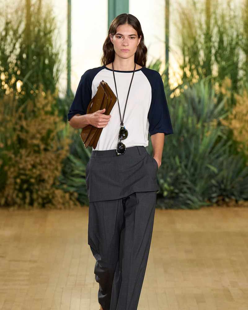 Modern casual look featuring a model in a white and navy raglan tee paired with relaxed grey trousers%2C accessorized with a brown clutch and sunglasses.
