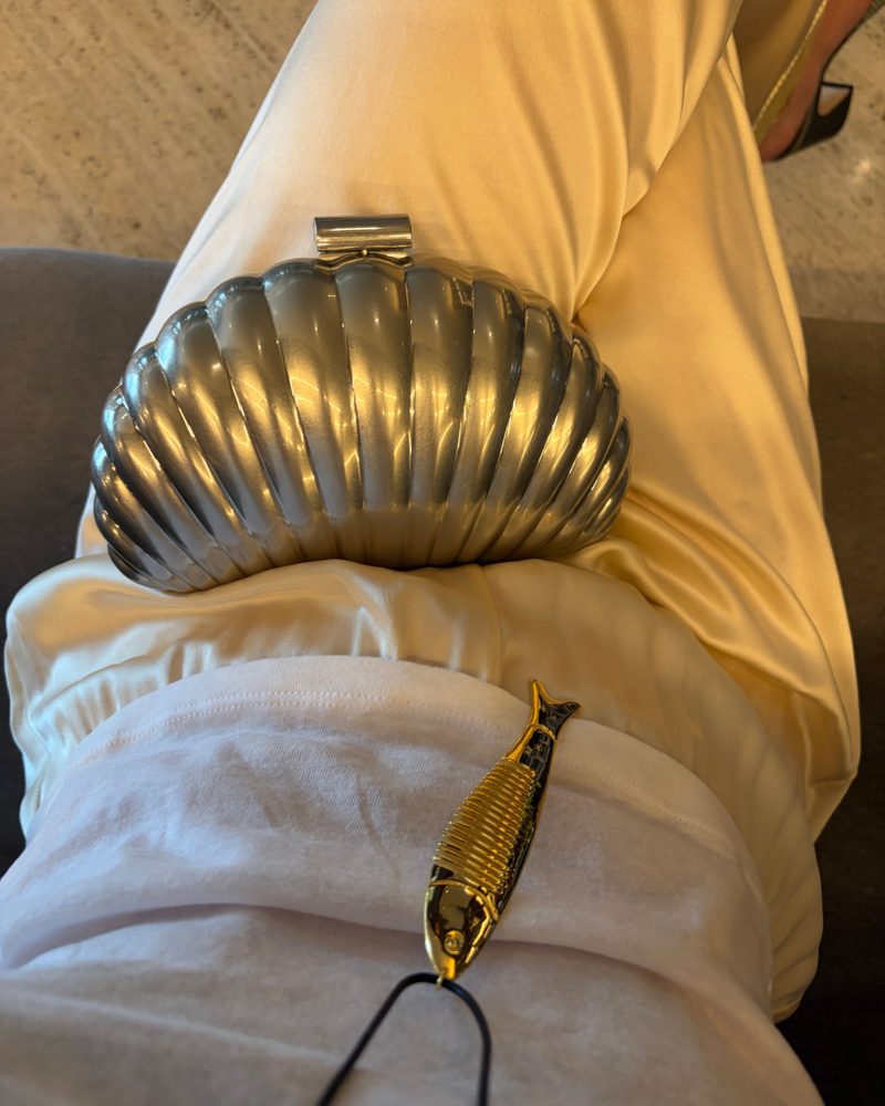 Silver metallic shell-shaped clutch paired with cream satin pants and a gold fish-shaped belt accessory for a chic%2C modern look.