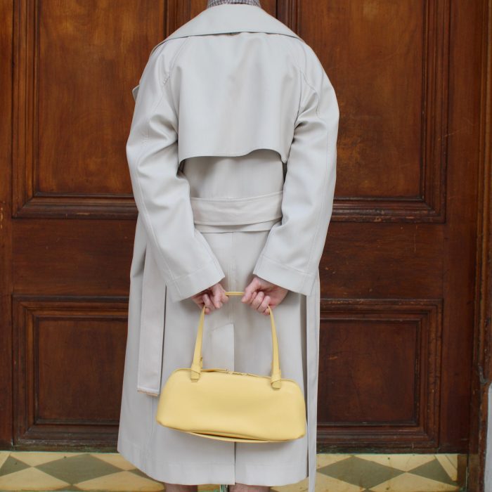 Back view of beige trench coat with yellow leather handbag and brown loafers, posed at a wooden doorway