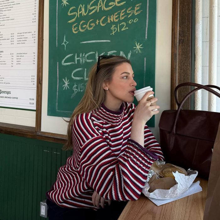 A person in a red and white striped sweater sips coffee at a cafe. A chalkboard menu lists prices; a bag and food on the table. Cozy, relaxed mood.